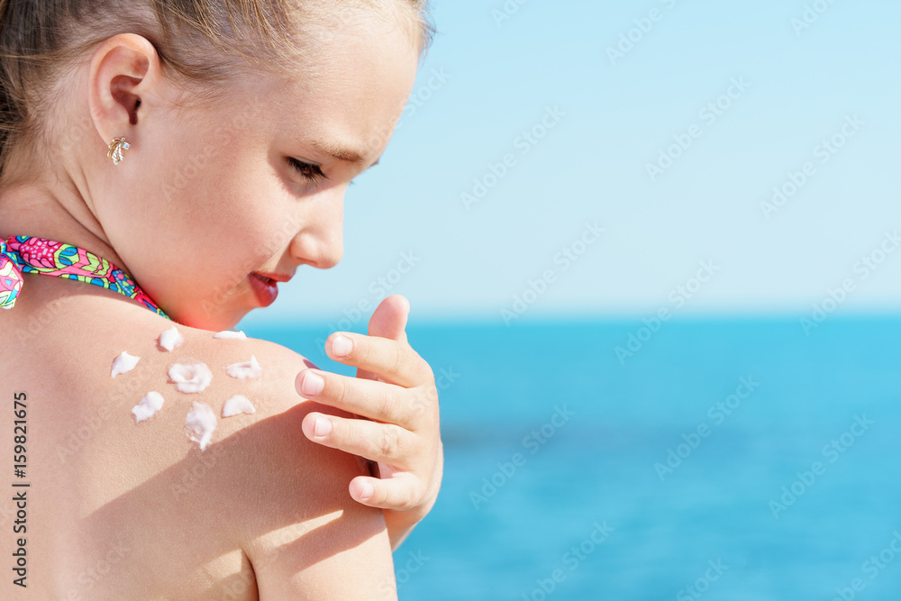 young girl applyng sun protector cream at her shoulder on the beach ...