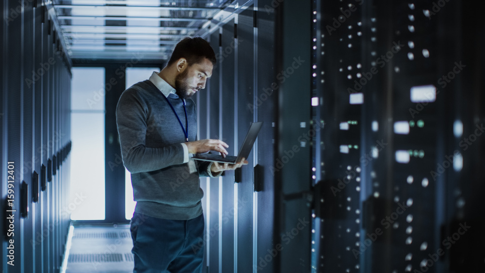 IT Technician Works on Laptop next to a Server Cabinet in Big Data ...