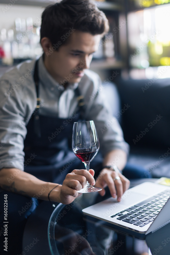 Portrait of senior sommelier sitting in front of laptop at wine cellar ...
