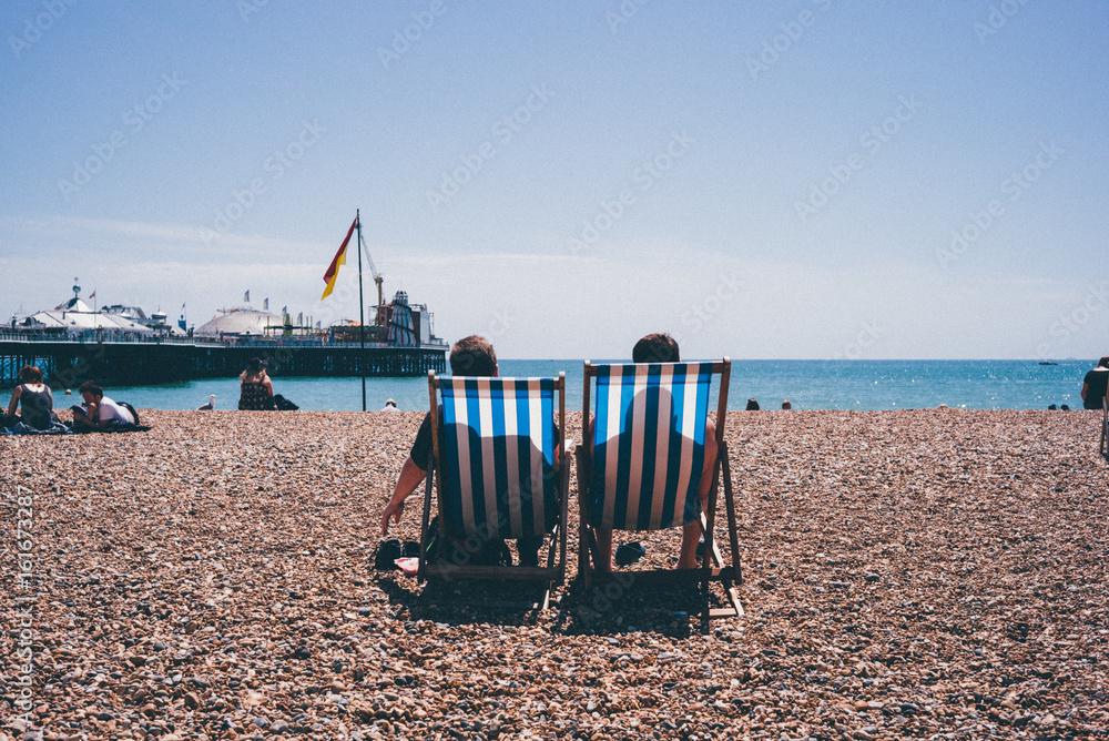 A classic British seaside scene. 2 people sat on deckchairs on stone ...