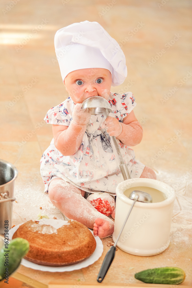 Cute liitle girl in chef's hat sitting on the kitchen floor soiled with ...