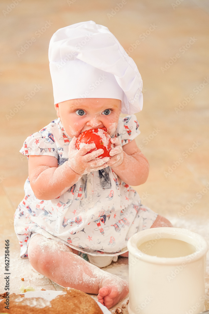 Cute liitle girl in chef's hat sitting on the kitchen floor soiled with ...