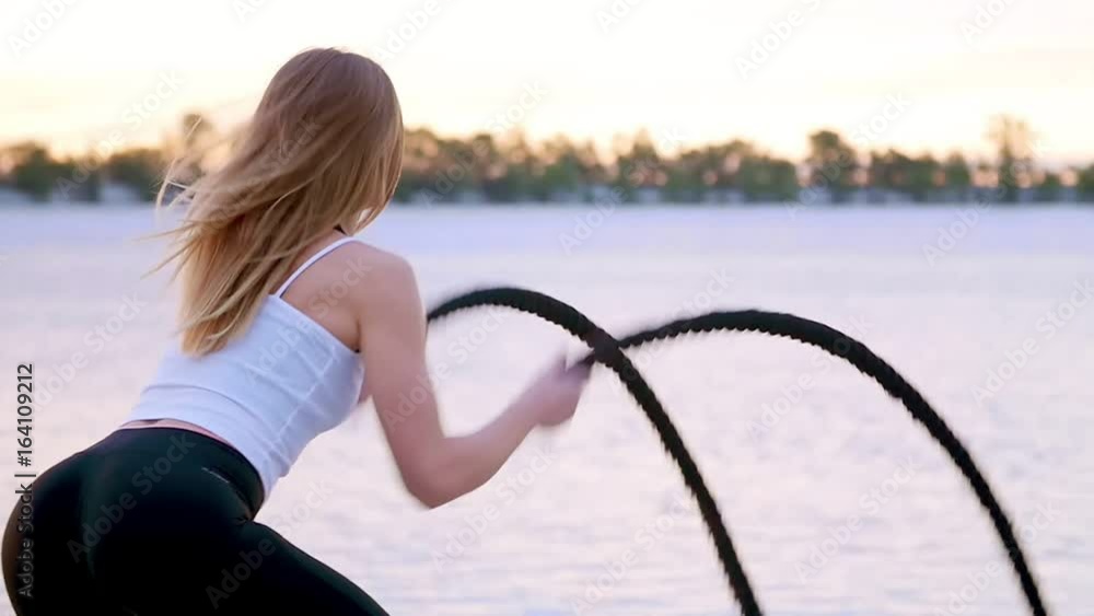 Young, athletic, blond woman, performs strength exercises with the help of a thick, sporty rope ...
