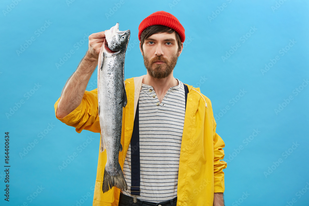Bearded young man angling big fish in pond, posing with it over blue ...
