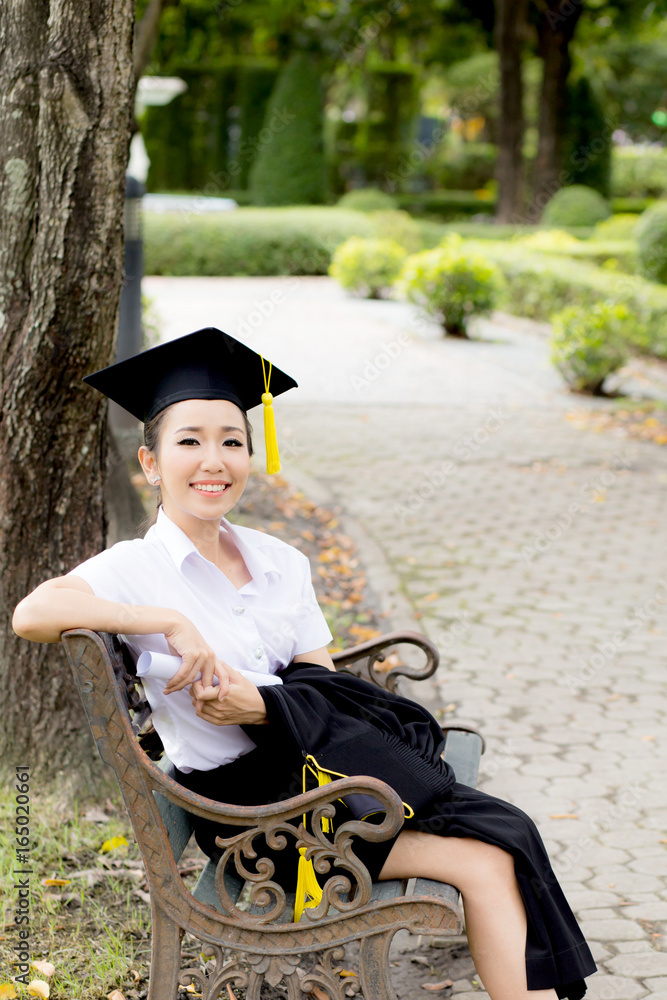 Joyful student celebrating his graduation seated on a bench in a park ...