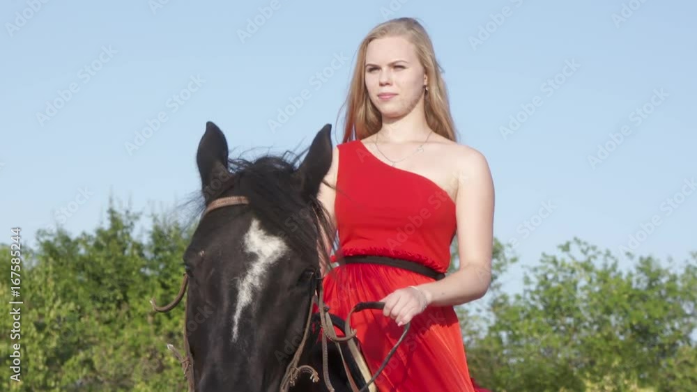 Young blonde girl wearing bright long red dress riding black horse ...