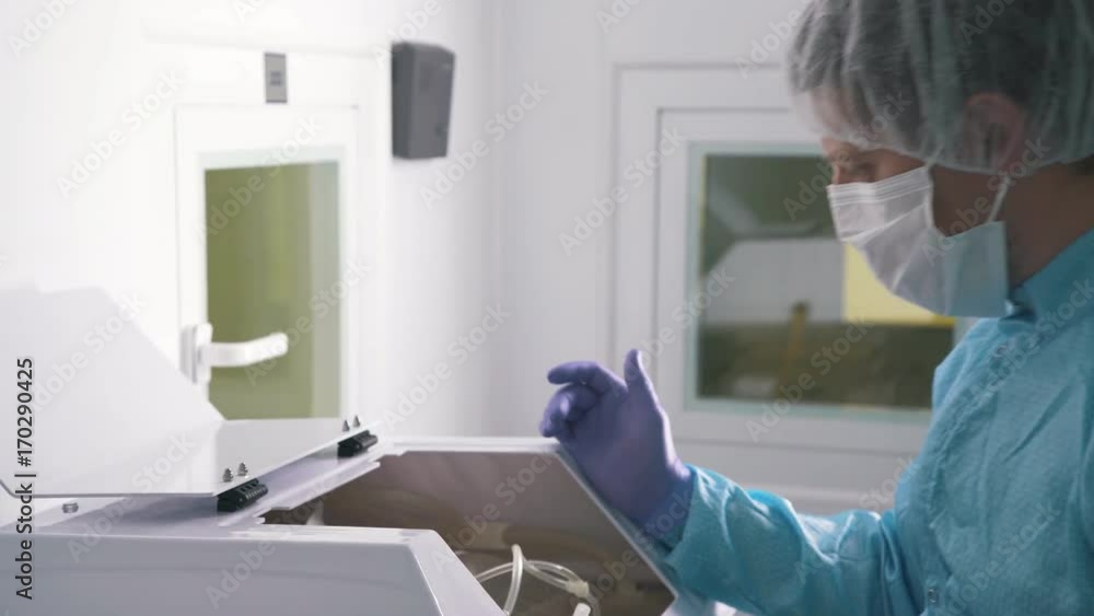 Scientist in a sterile suit (cap, face mask, latex gloves) makes ...