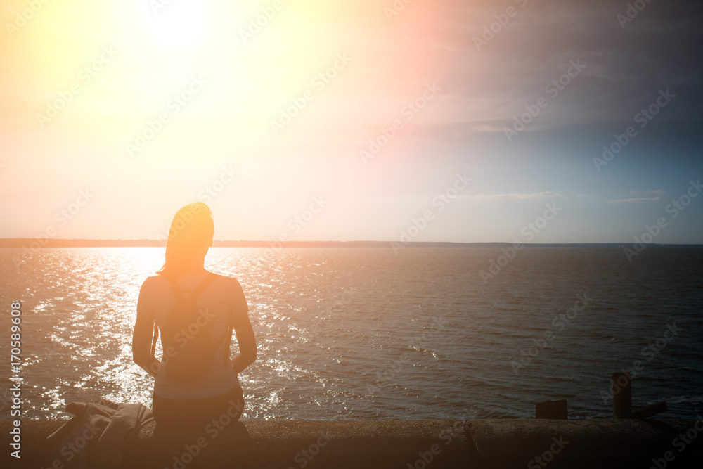 Girl sits on the stone embankment of coast sea and looks at the sun reflects on the surface of ...