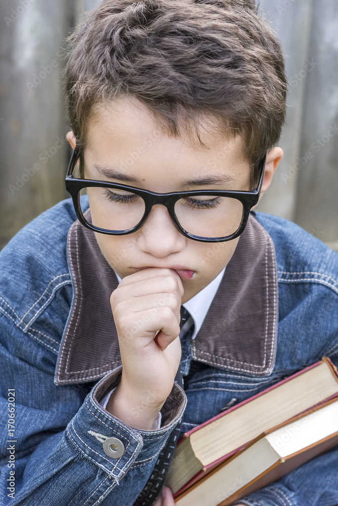 Preteen boy with black glasses holding books and leaning face on hand ...