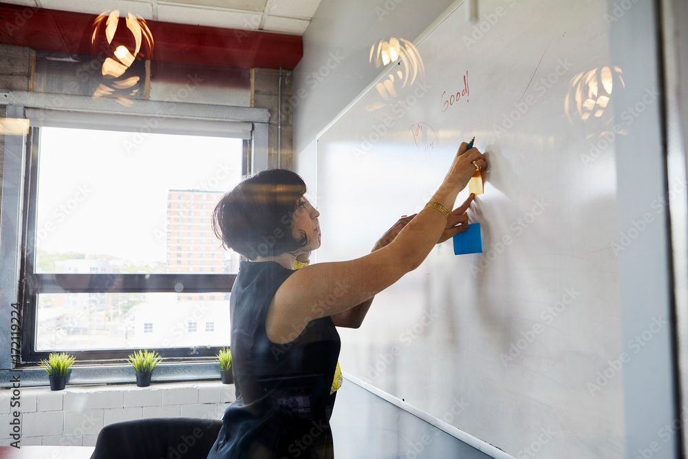 Two women in office, brainstorming, sticking sticky notes on whiteboard ...