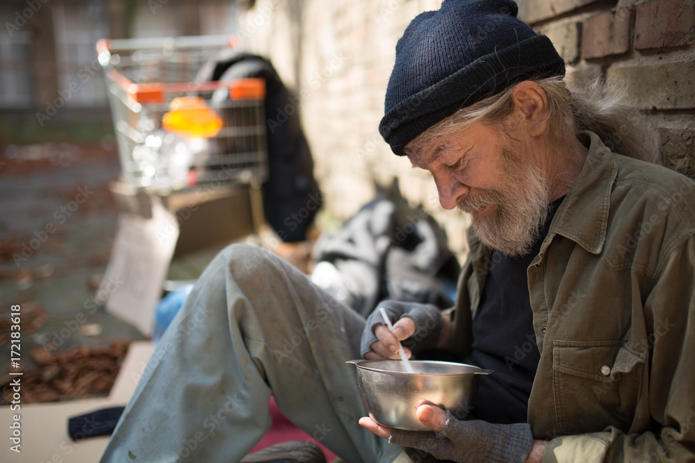 Close up view of tramp sitting by the brick wall, eating. Homeless man ...