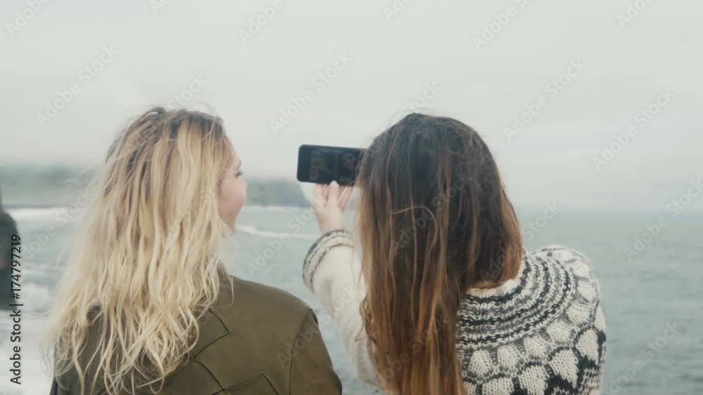 Back view of two young women taking selfie photos on the shore of the ...