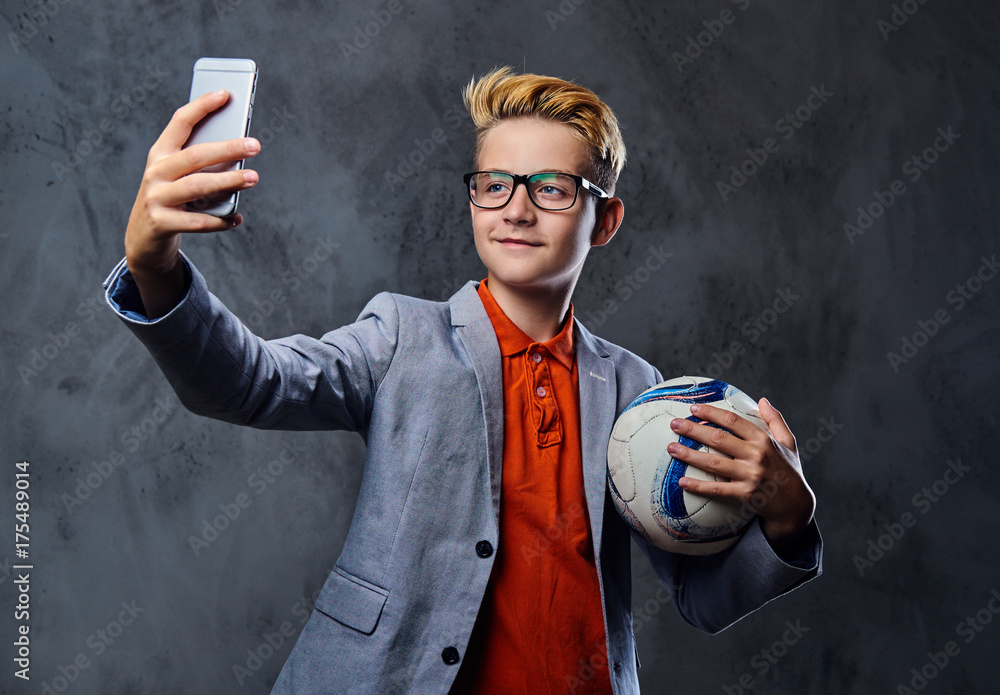 A boy in eyeglasses holds soccer ball and making selfie on a smartphone ...