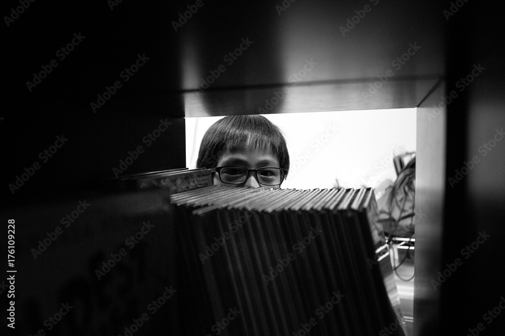 Young boy with glasses peering through a bookshelf with books inside a ...