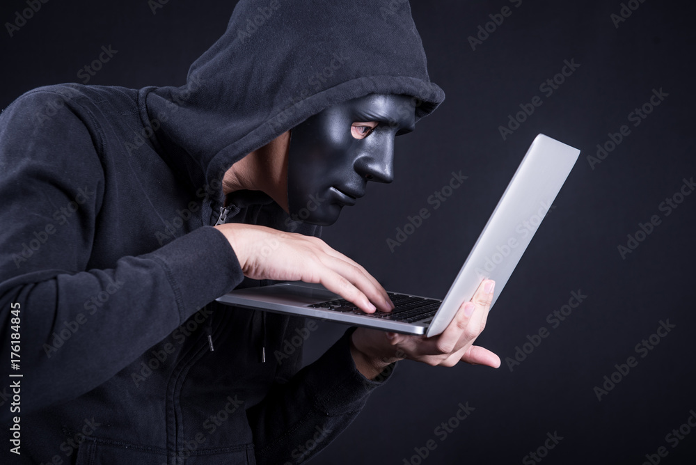 Anonymous male hacker using black mask to cover his face carrying laptop computer. Internet ...