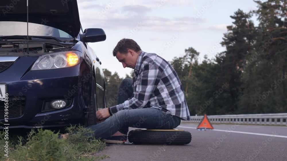 Caucasian man changing a tire sitting on stepney near car on the ...