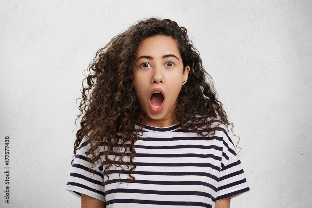 Surprised excited female with curly hair stares at camera, opens mouth ...