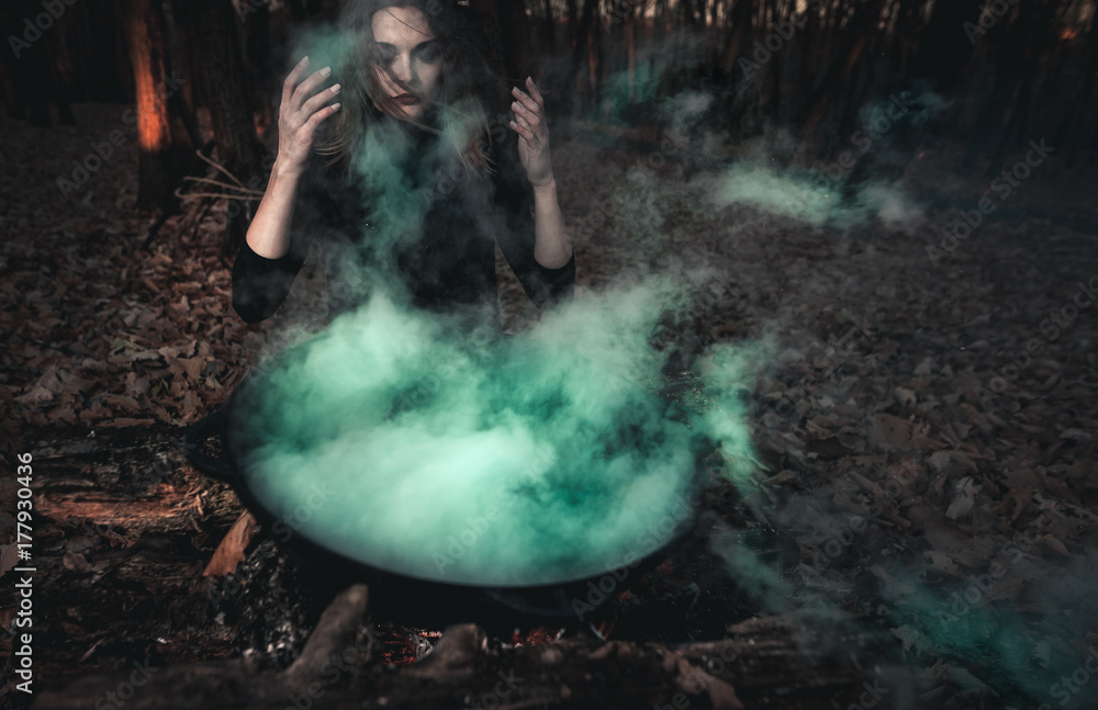 A beautiful girl witch cooking an infernal potion in a cauldron at the ...