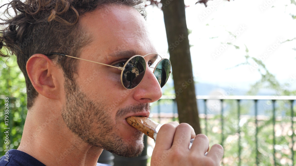 Handsome young man eating cereal bar, looking away sitting on terrace ...