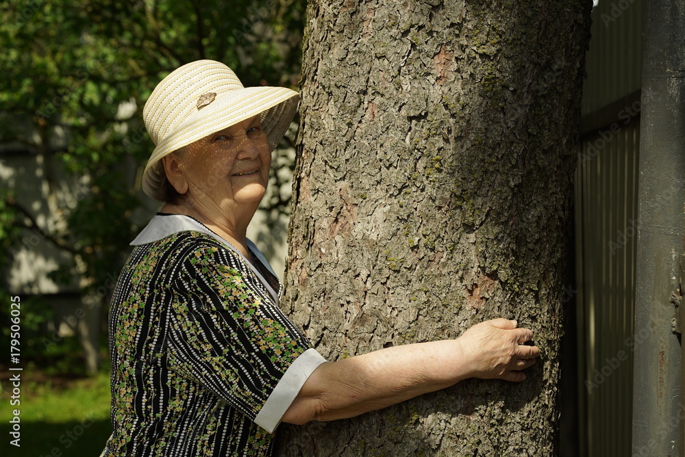 Elderly mature woman aged 80s dressed in white hat hugging tree trunk ...