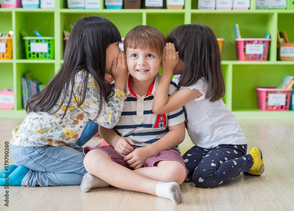 Two girl kids whisper secret at ear of boy in library at kindergarten ...