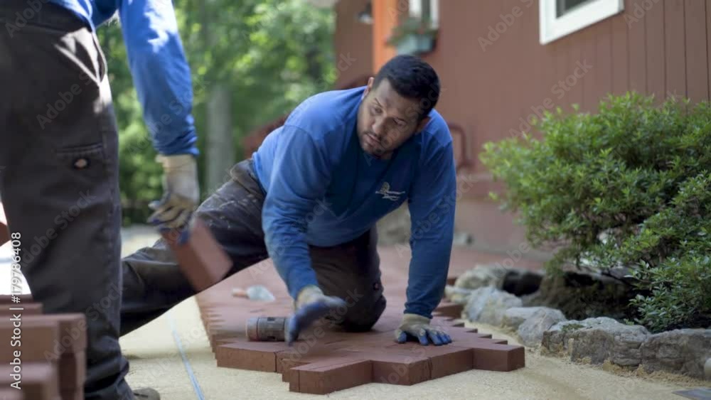 One man hands red bricks to another man who is putting the brick pavers ...
