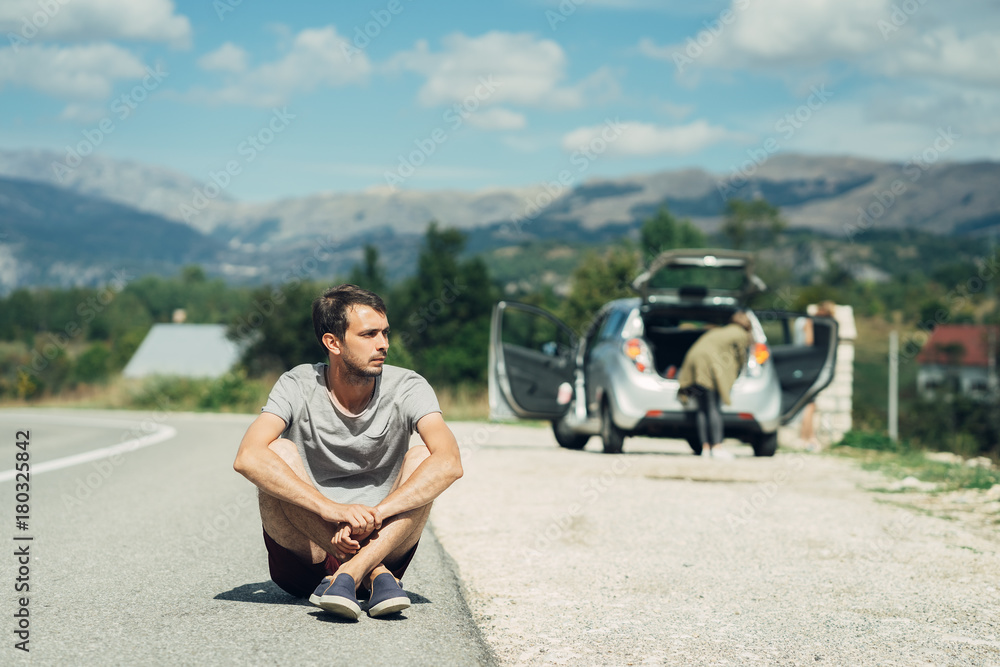 Young man is sitting in front of car on empty road. Road trip stop ...