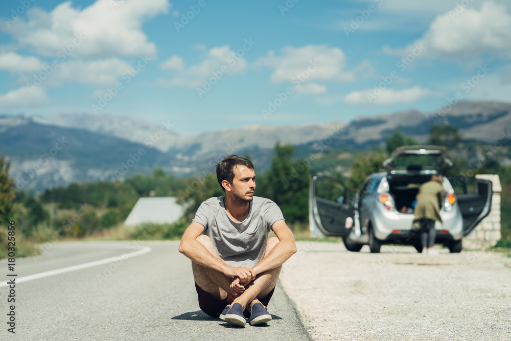 Young man is sitting in front of car on empty road. Road trip stop ...