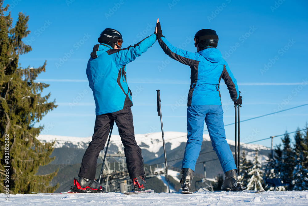 Pair giving high five to each other, smiling looking standing with skis ...