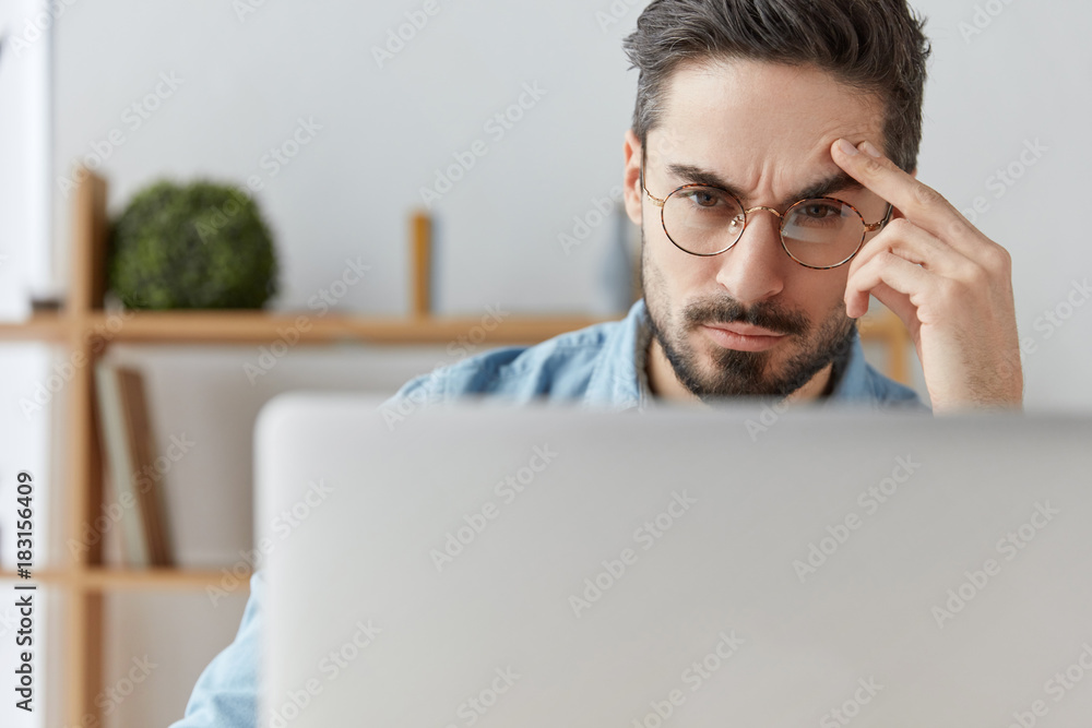Concentrated bearded man looks attentively at screen of laptop computer ...