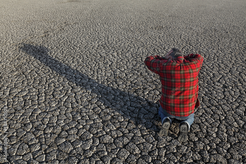 Desperate man kneeling and holding his head in his hands at dry cracked ...