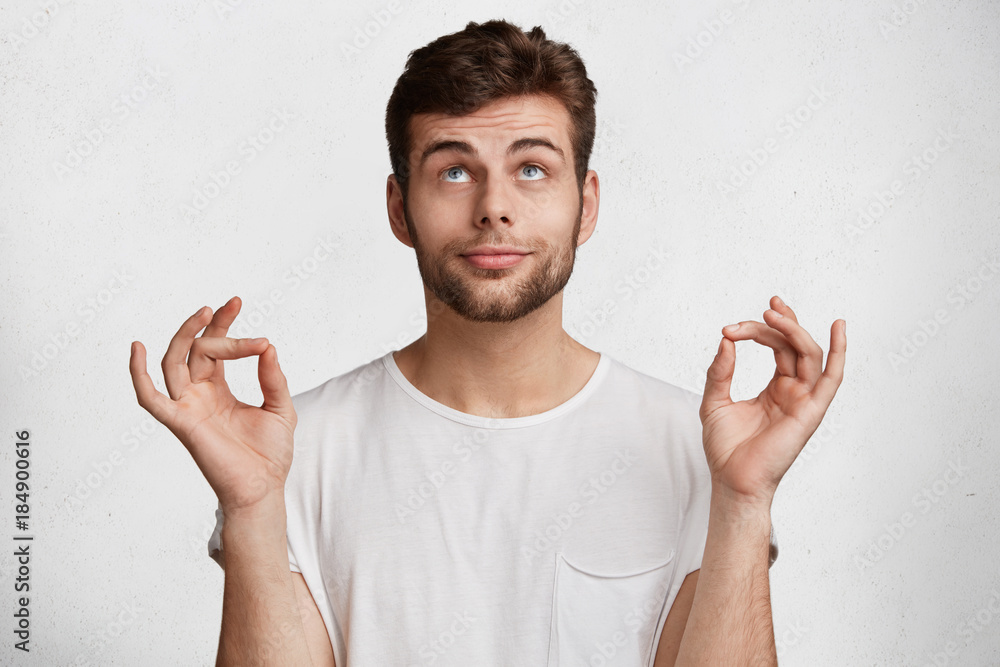 Young European male has peaceful expression, keeps hands in mudra ...