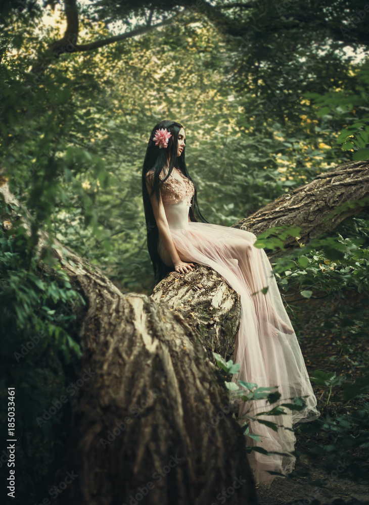 A fragile girl, in a transparent dress, sits on a branch. A forest ...