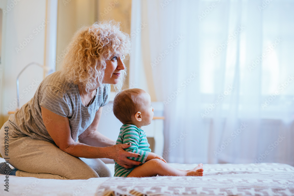 Blond curly female nanny with infant baby boy sitting on the bed in ...