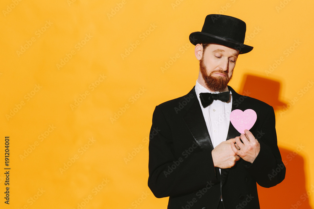 Sad man in tux and top hat holding a pink heart isolated on orange ...