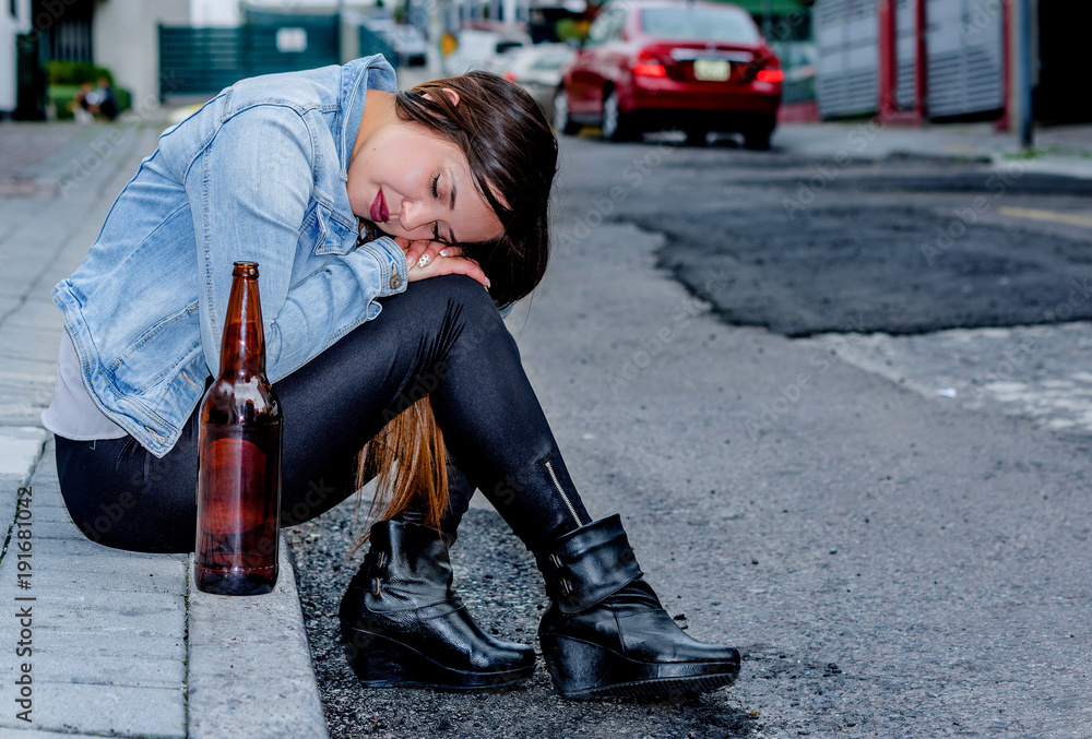 Beautiful young drunk woman sitting in a sidewalk with bottle of beer ...