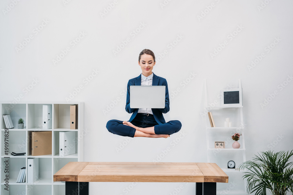 smiling young businesswoman using laptop while levitating in office ...