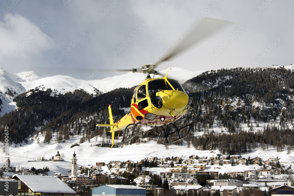A yellow helicopter flying up in the air over the small town in Engadin ...