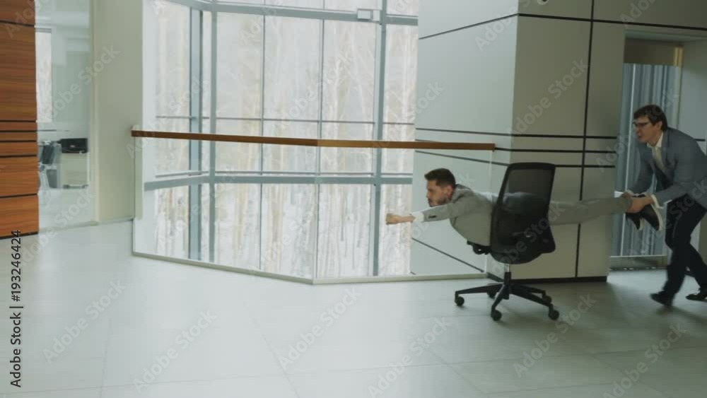 Slow motion pan shot of two funny businessmen riding office chair while ...