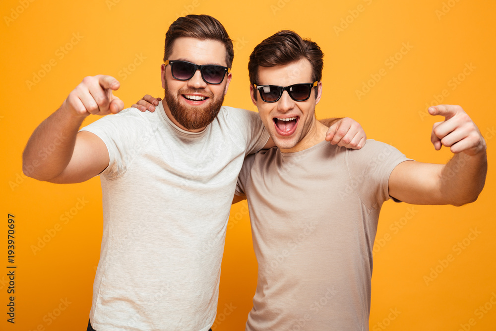 Two happy men in casual t-shirts and sunglasses hugging and pointing ...