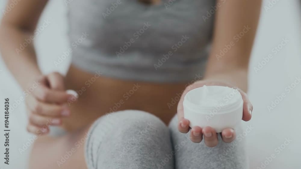 A young girl applies a body cream on her leg. A woman holds a jar of ...