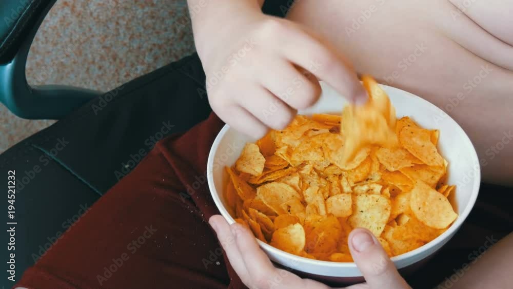Fat teenager with fat folds on his body is eating potato chips with his ...