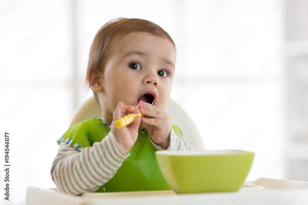 Baby child eats food himself with spoon. Portrait of happy kid boy in ...