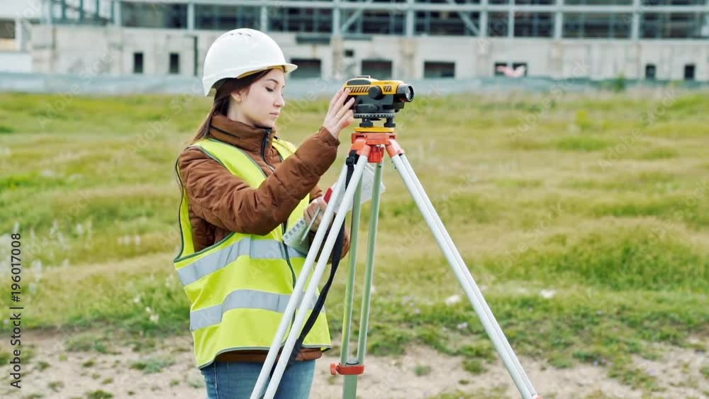 A young woman surveyor in work clothes and helmet adjusts the equipment ...