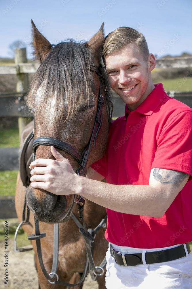Handsome, smiling horse rider standing next to and petting his horse ...