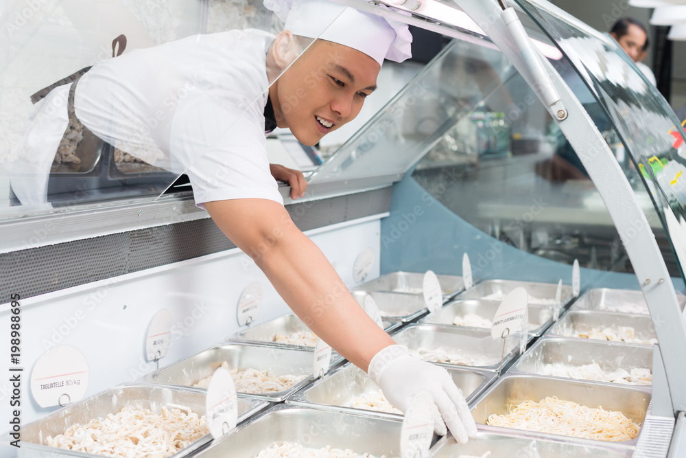 Asian chef placing food in trays into a glass counter in a delicatessen ...