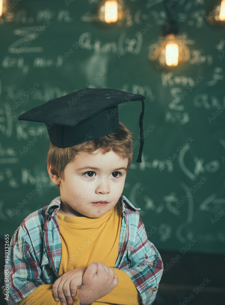 Smart child in graduate cap on serious face, shy, holds hands crossed ...