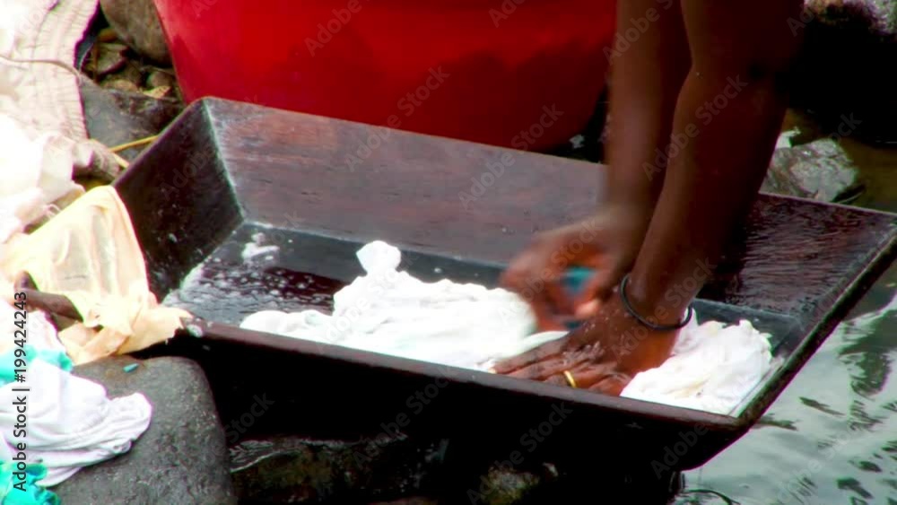 African woman uses a small wooden bin and a rock to wash clothing in a ...