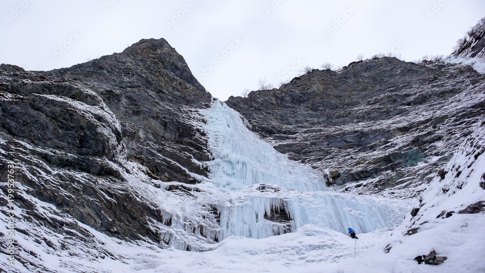 male ice climber stands at the headwall of a long and steep waterfall ...