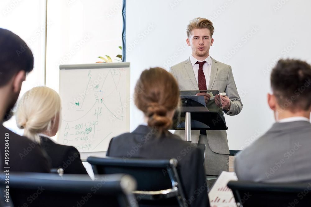 Portrait of young businessman standing by podium while giving speech at ...